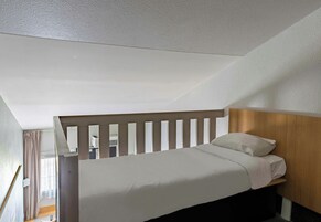Uma cama de solteiro com cabeceira de madeira num quarto com teto inclinado e janela com cortinas.