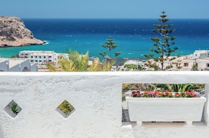 Vista costeira com um prédio branco, uma varanda florida e mar azul límpido.