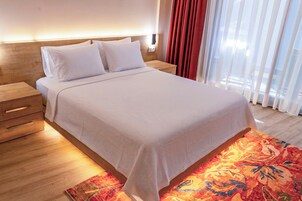 Cama bem arrumada com lençóis brancos, mesa de cabeceira de madeira com gavetas e tapete colorido no chão.