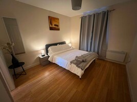 Quarto com cama, mesas de cabeceira, um radiador, janela com cortinas e um quadro na parede.