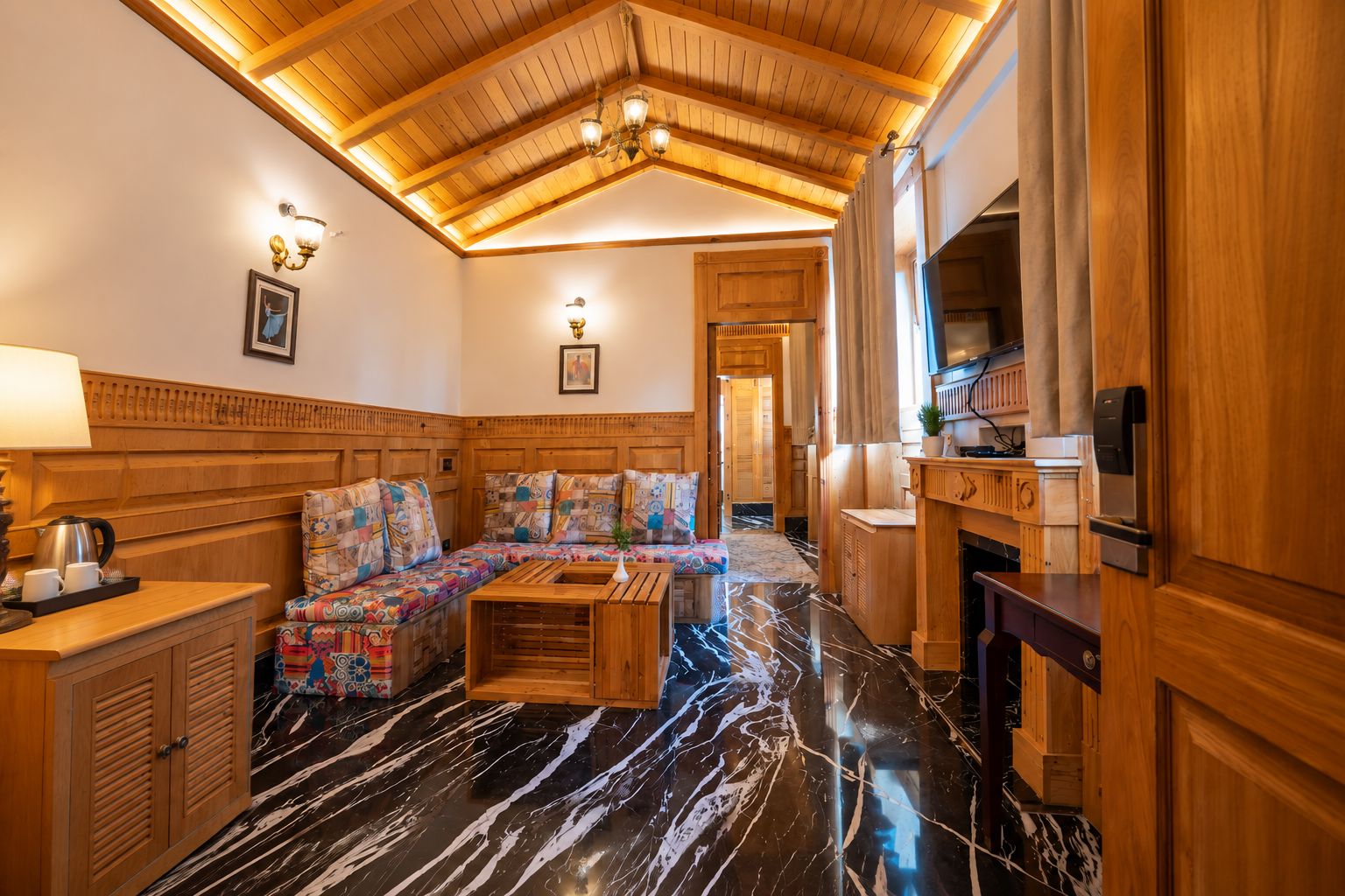 Quarto aconchegante com teto de madeira, um sofá com almofadas coloridas, uma mesa de centro de madeira e uma área de cozinha com armários de madeira e uma geladeira.