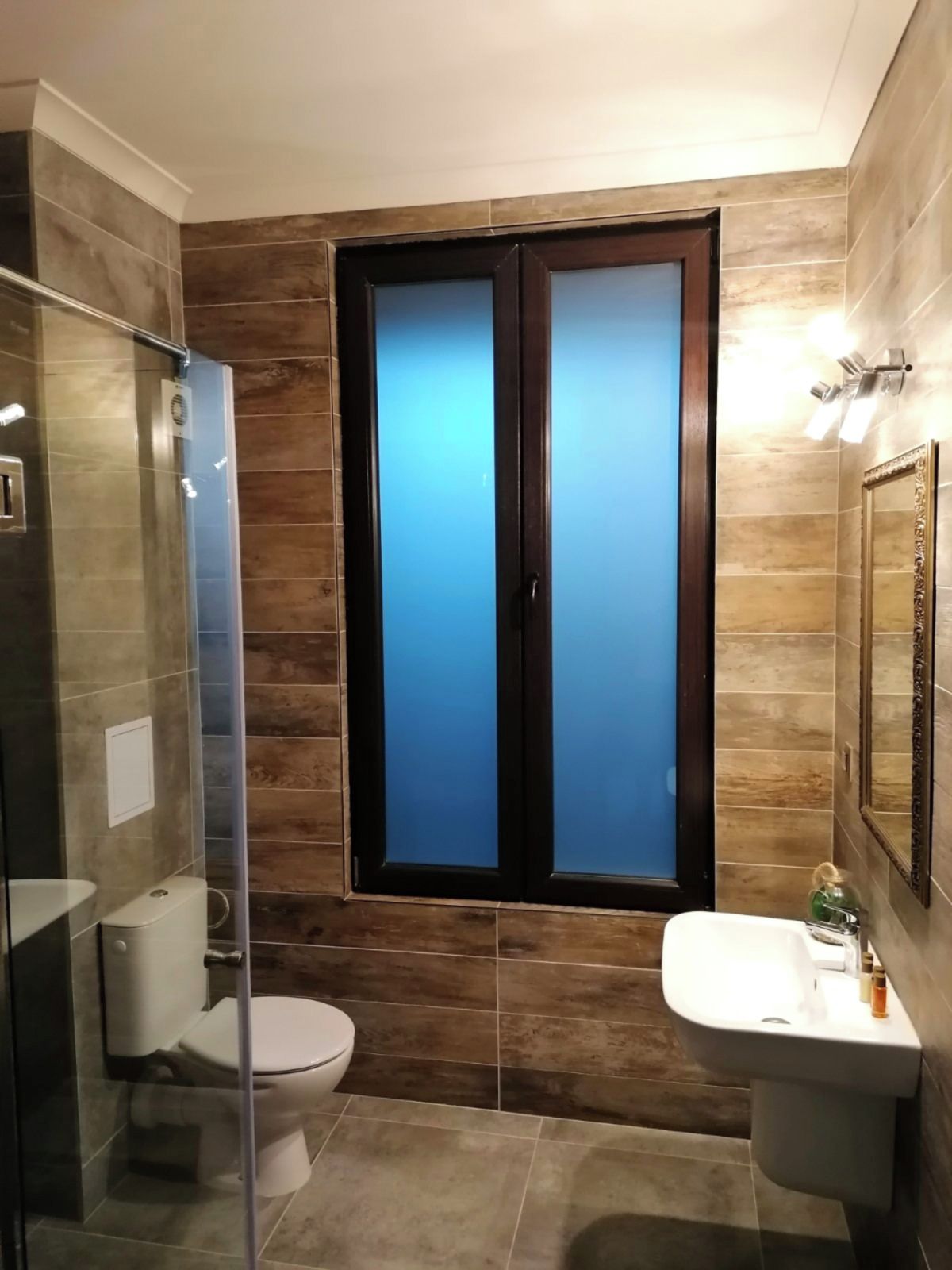 Casa de banho moderna com cabine de duche em vidro, sanita, lavatório com espelho e uma janela ampla com painéis de vidro jateado.