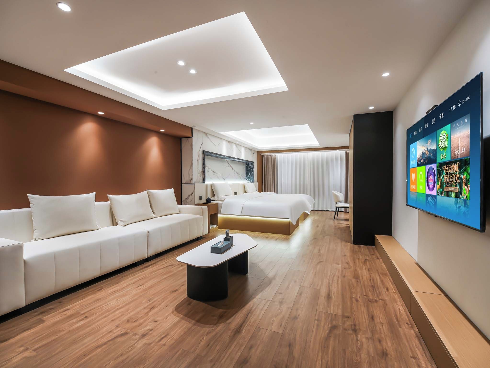 Quarto de hotel moderno com uma cama grande, um sofá, uma mesa de centro e uma TV fixada na parede exibindo várias telas.