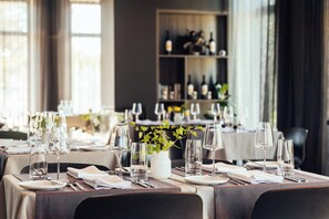 Ambiente de restaurante com mesas preparadas para refeições, com copos de vidro transparente e um vaso com folhagem verde.