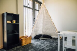 Um quarto acolhedor com uma tenda de brincar trançada, um cesto de vime, um sofá preto e uma mesa branca.