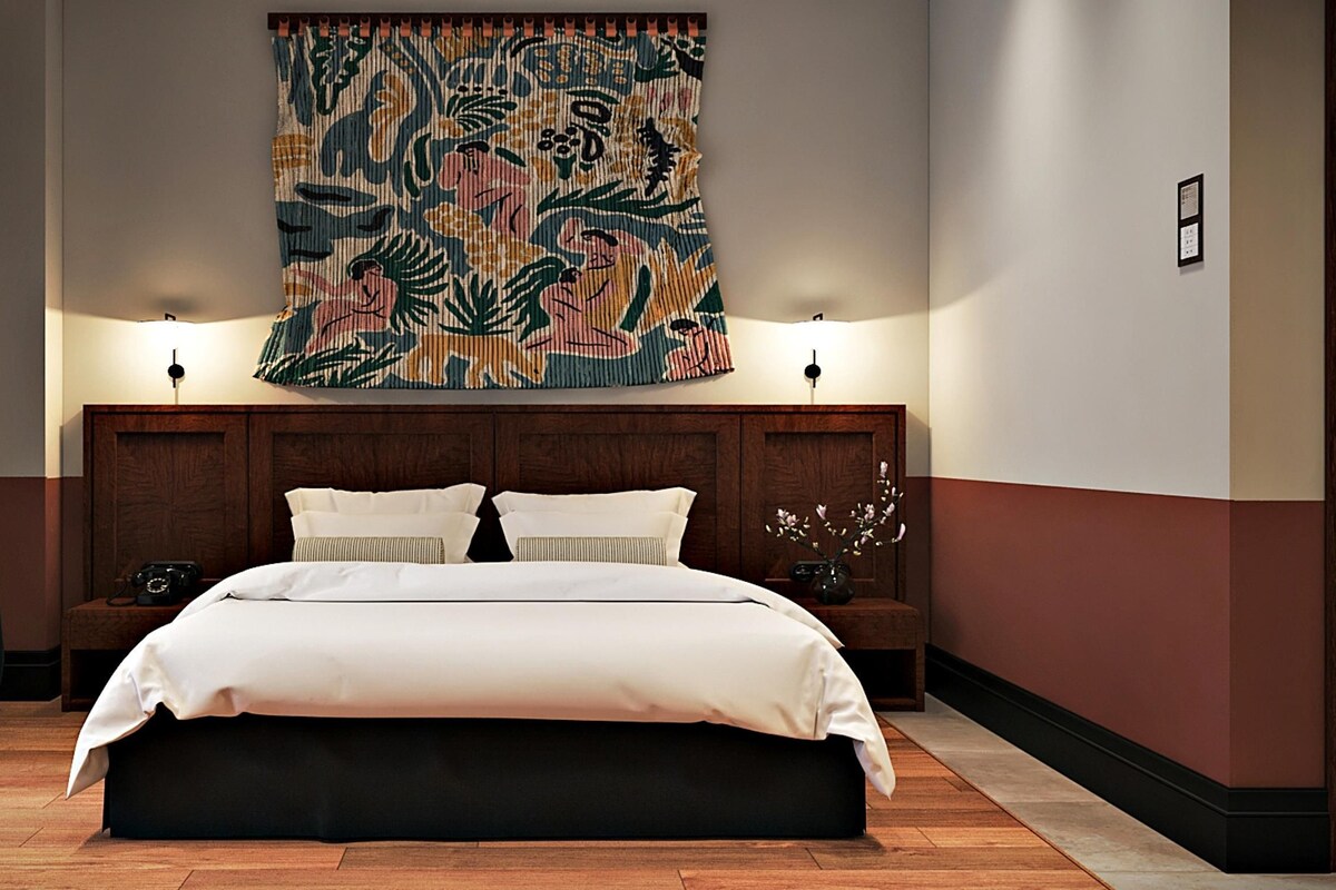 Quarto de hotel com uma cama grande, cabeceira com tapeçaria, abajures e piso de madeira.