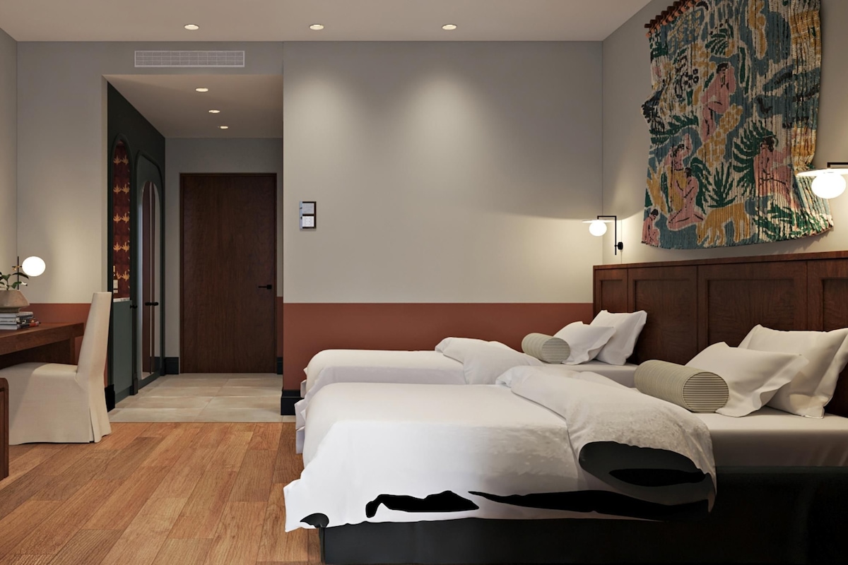 Quarto de hotel com uma cama, mesas de cabeceira, uma escrivaninha com televisão e uma luminária, e um grande tapeçário na parede.