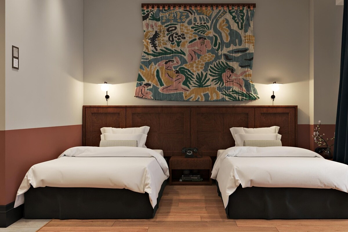 Quarto de hotel com duas camas, uma tapeçaria grande e uma janela com cortinas.