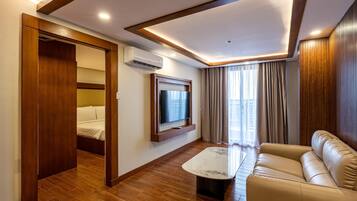 Quarto de hotel com piso de madeira, sofá, televisão fixada na parede e cama com guarda-roupa de madeira.