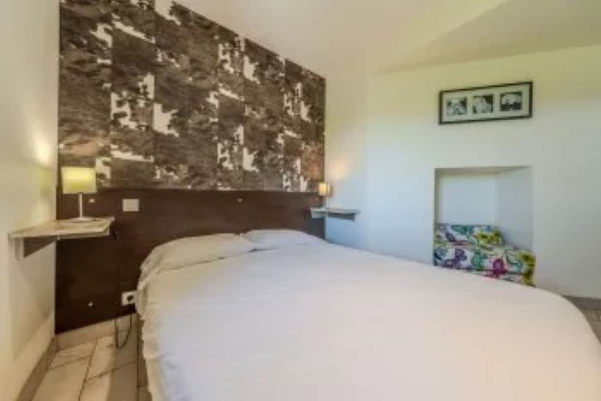 Quarto com cama, mesa de cabeceira, luminária, prateleira na parede e quadro emoldurado.