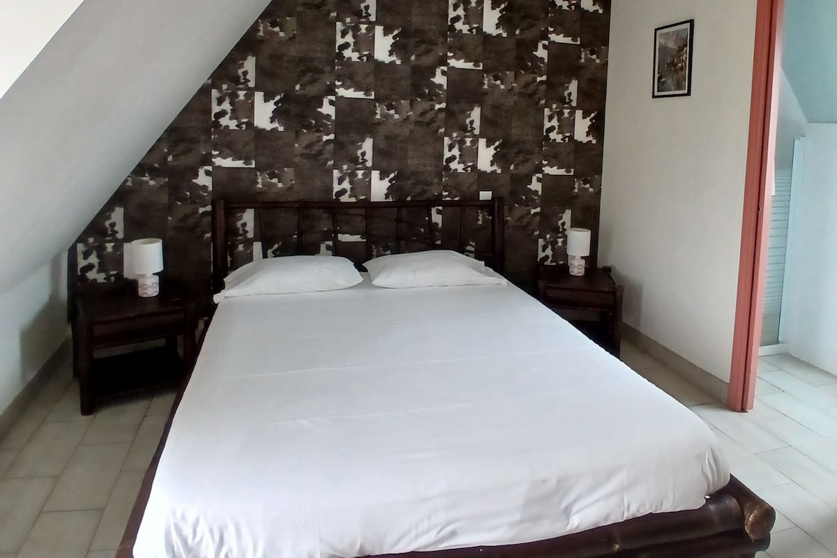 Quarto com teto inclinado, cama com roupa de cama branca, cabeceira de madeira, duas mesas de cabeceira com luminárias e uma parede com padrão de camuflagem.