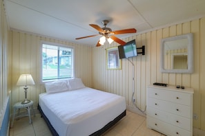 Quarto com cama, ventoinha no teto, TV fixada na parede, cômoda e janela com persianas.