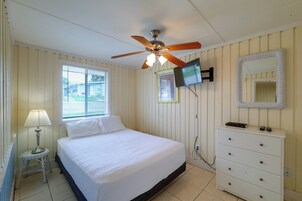 Quarto com cama, ventoinha no teto, TV fixada na parede, cômoda e janela com persianas.