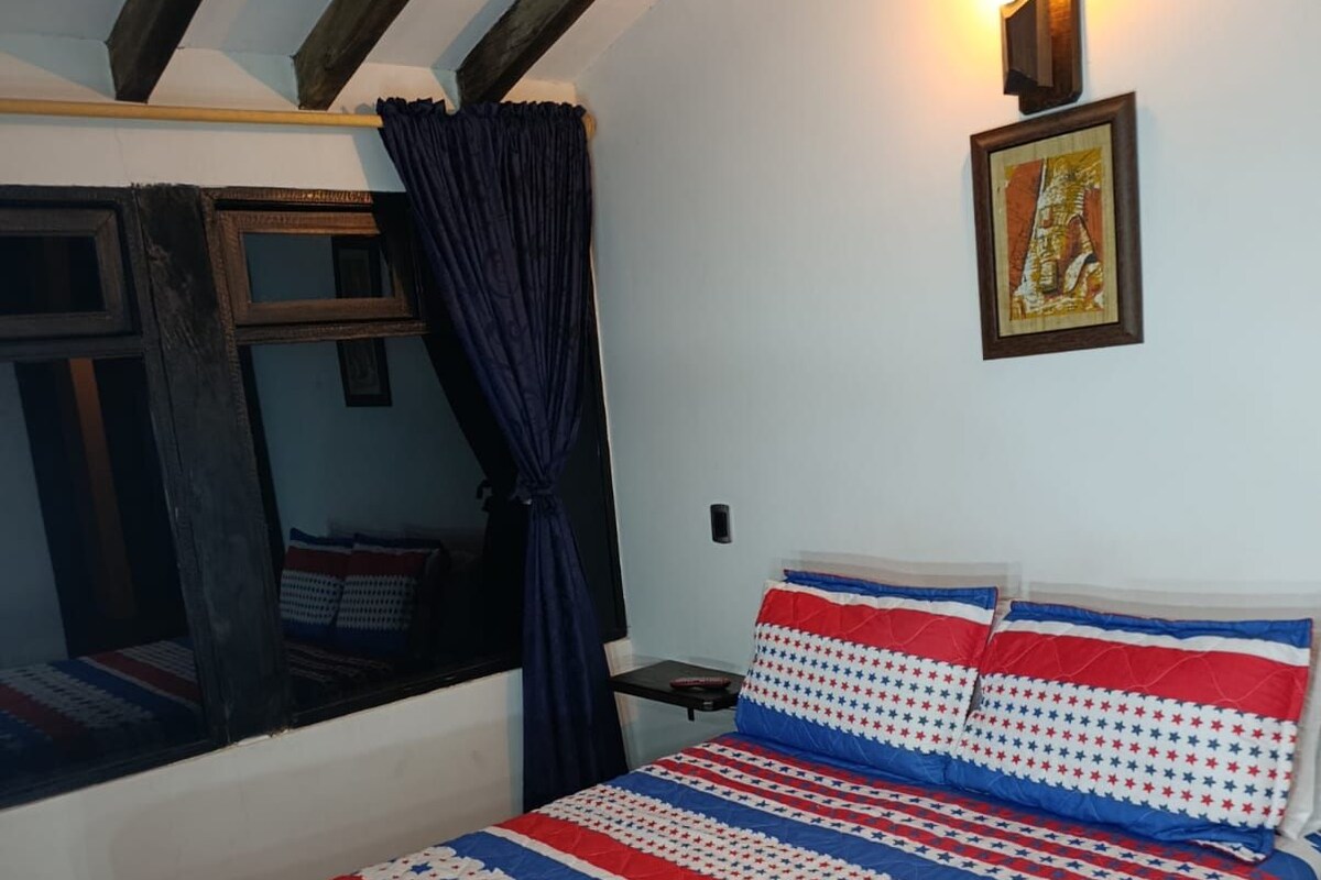 Quarto com cama, mesa de cabeceira, luminária na parede, janela com cortinas e um quadro emoldurado.