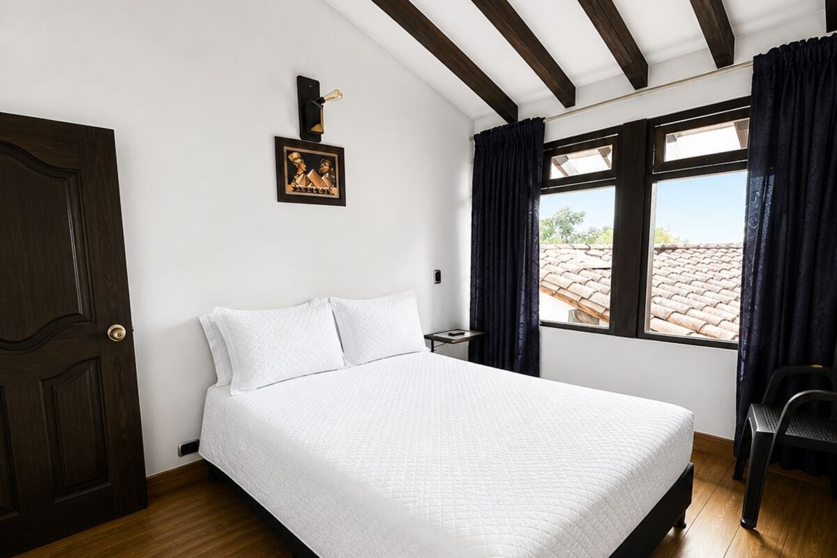 Quarto de hotel com uma cama grande, piso de madeira, porta escura, janela com cortinas e uma pequena mesa de cabeceira.