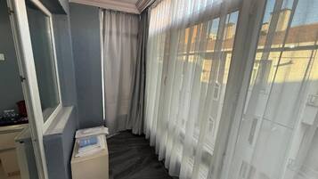 Quarto com uma janela grande, piso de madeira e uma pequena mesa com objetos sobre ela.