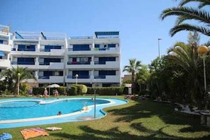 Hotel com vários andares, piscina, palmeiras e vegetação.