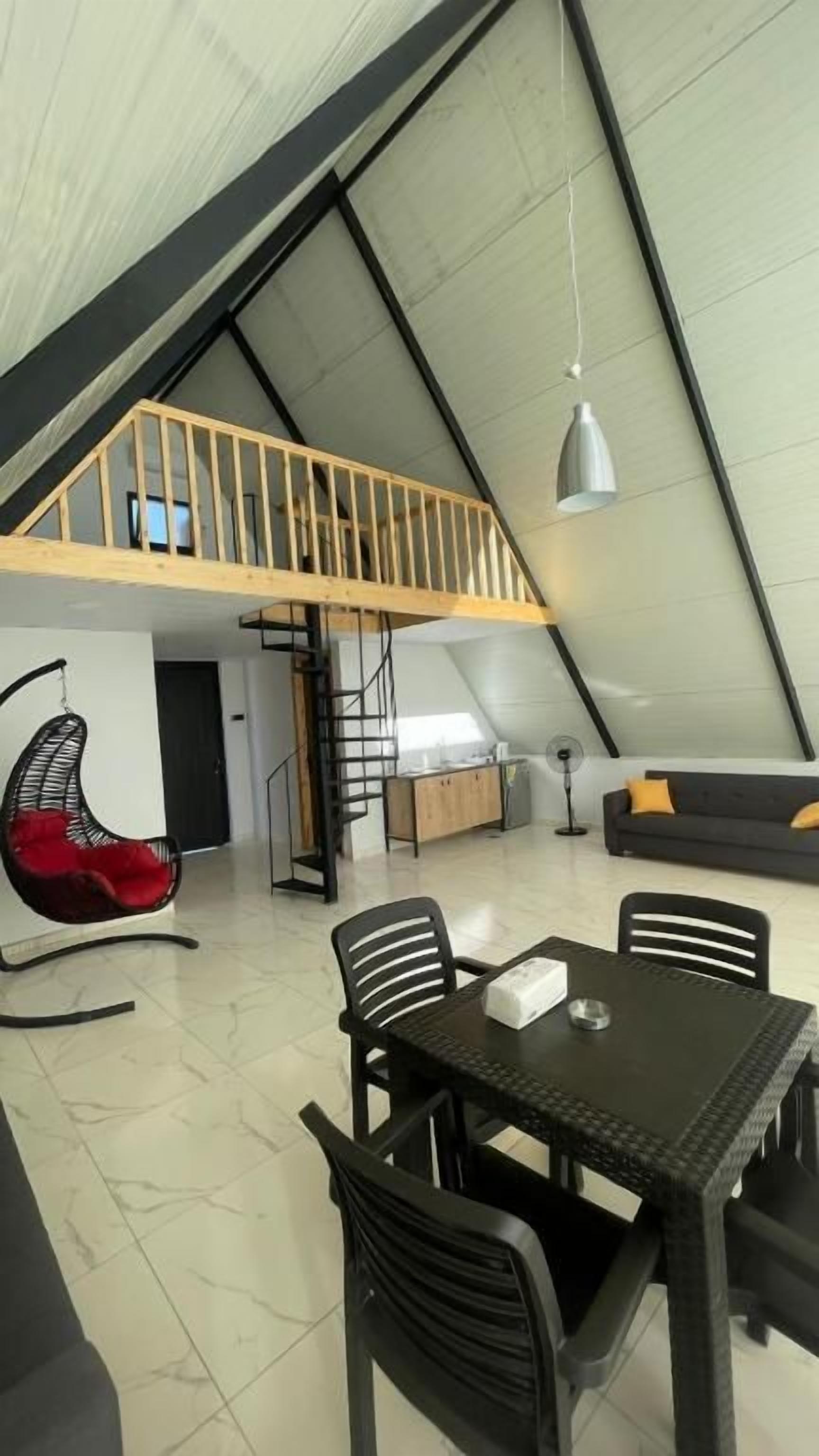 Um amplo loft com pé-alto, área de refeições com mesa e cadeiras, uma cadeira suspensa vermelha e uma escada que dá acesso a um mezanino com espaço para dormir.