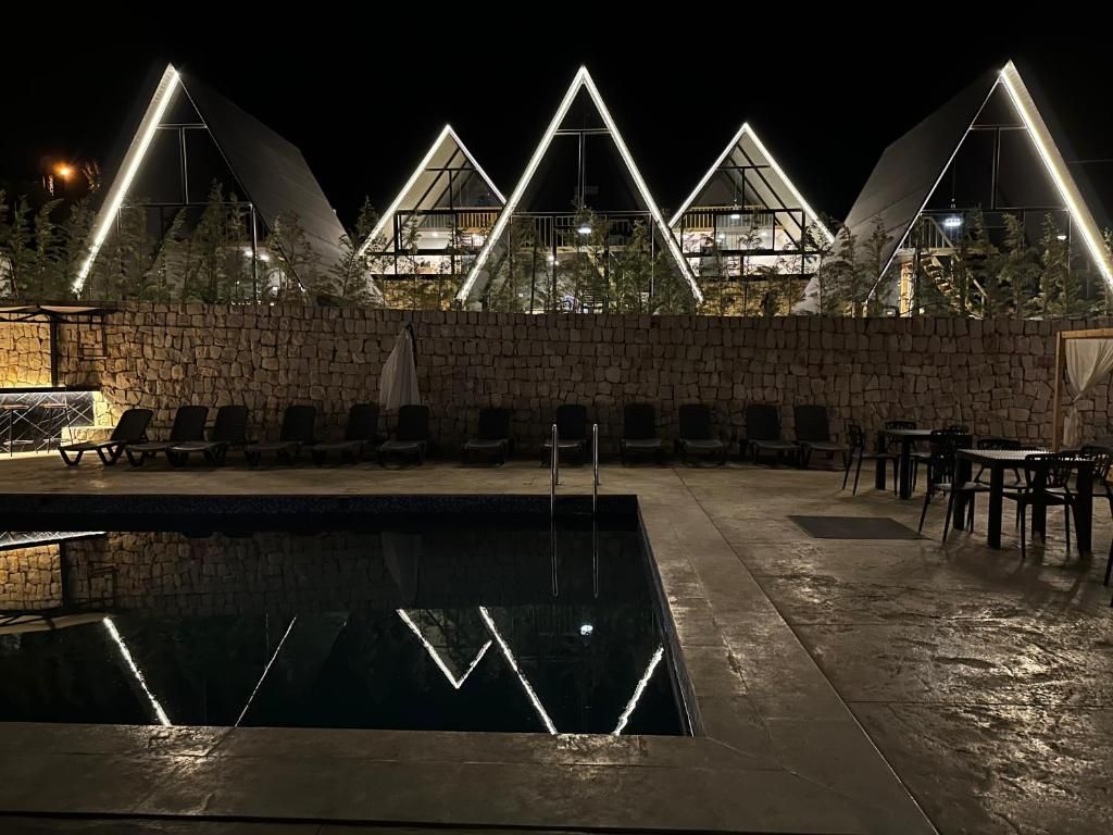 Piscina com espreguiçadeiras e mesas, cercada por um muro de pedra e estruturas de iluminação triangulares.