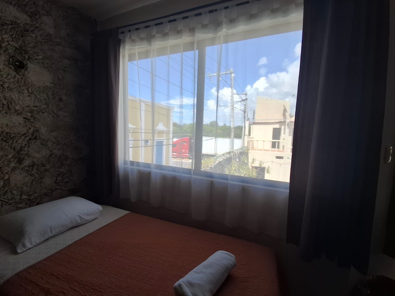 Quarto com cama, janela com cortinas e vista para prédios.
