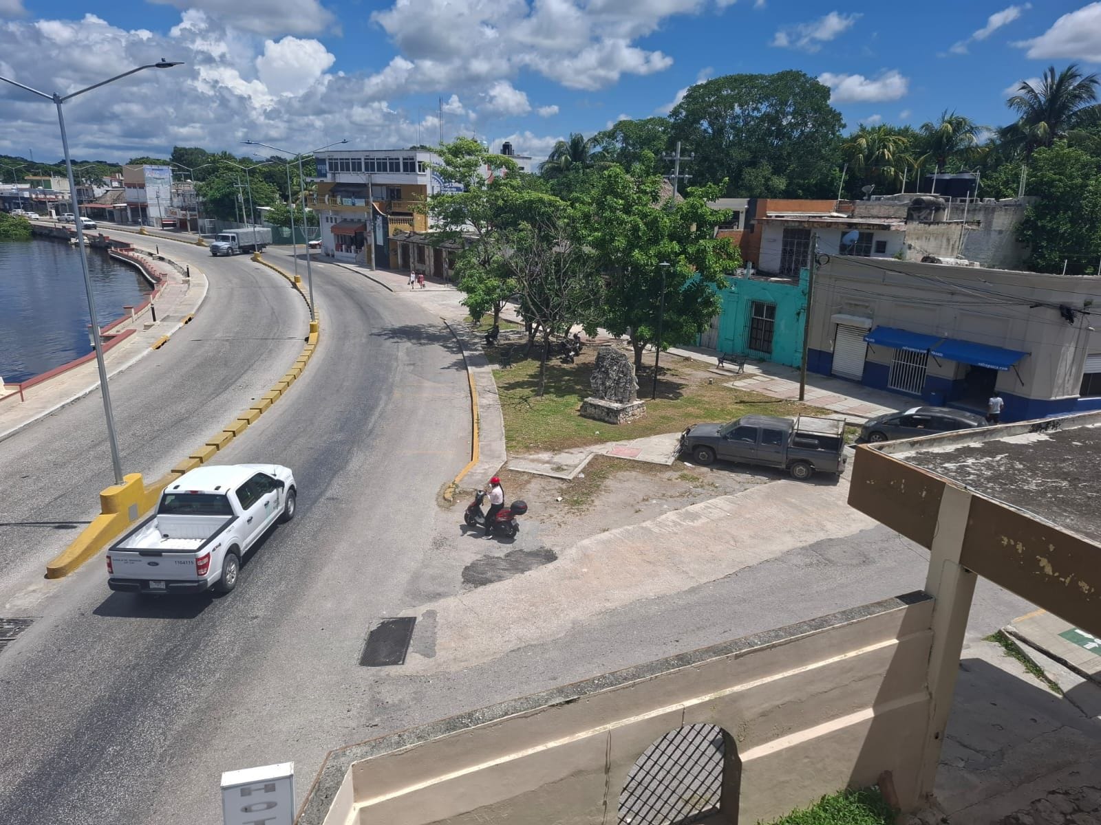 Vista da rua com um carro branco, uma moto e um carro estacionado. Há prédios, árvores e um céu claro.