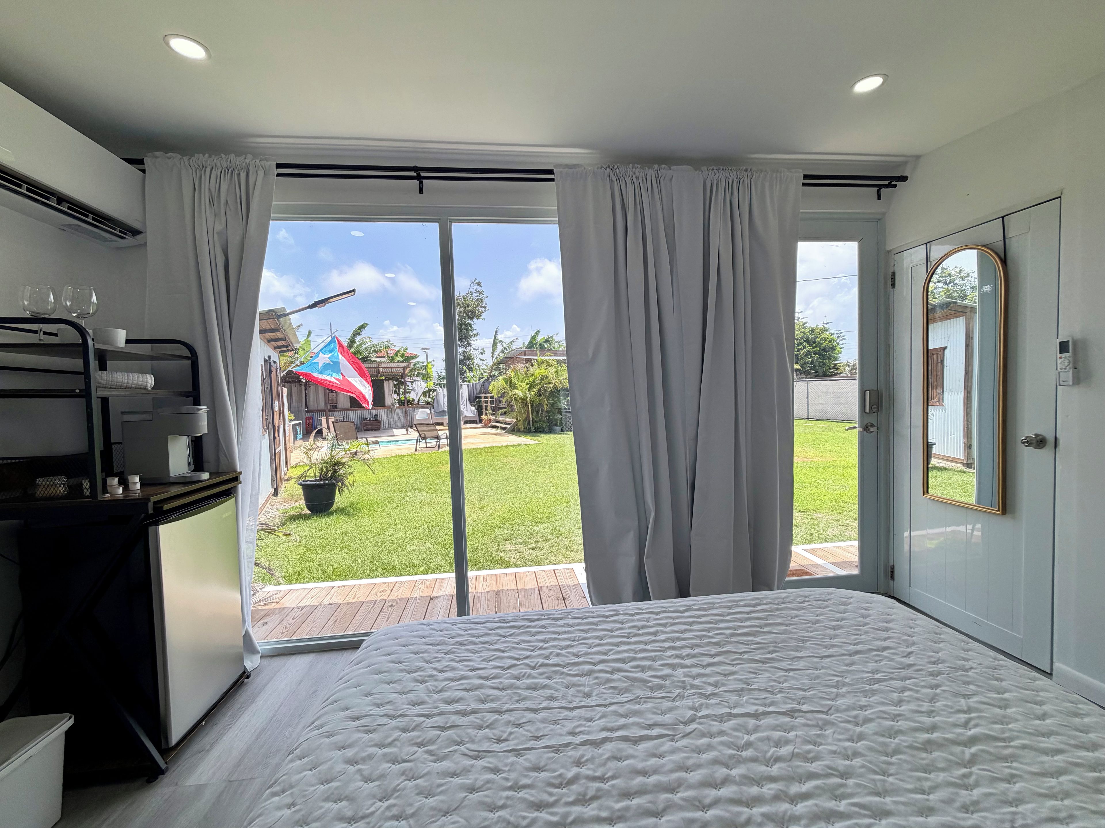 Quarto com cama grande, escrivaninha com computador e vista para um quintal com piscina e uma bandeira.