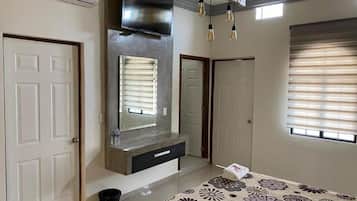 Quarto com cama, penteadeira, espelho, TV fixada na parede e janela com persianas.