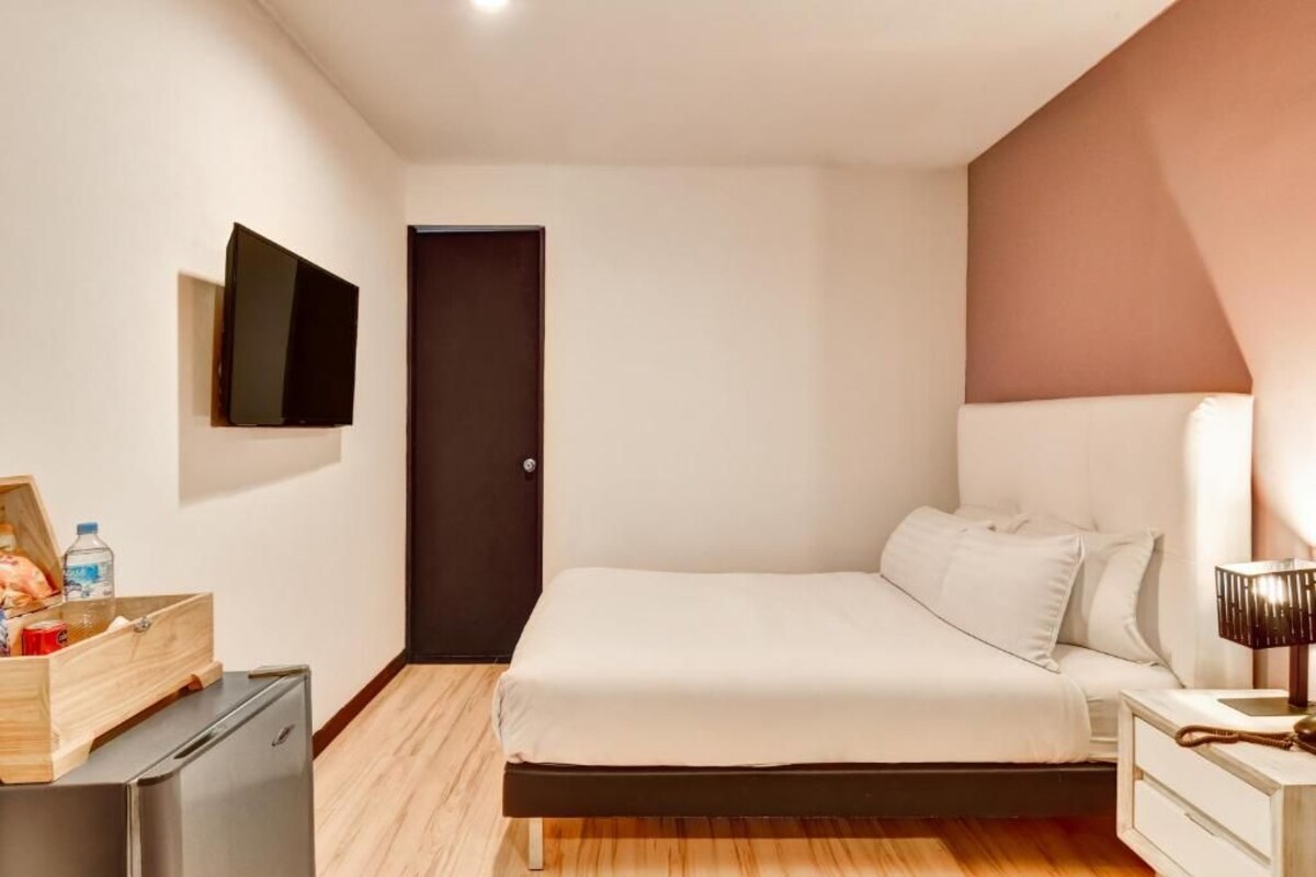 Quarto com cama, televisão de tela plana, cômoda de madeira, porta e mesa de cabeceira com luminária.