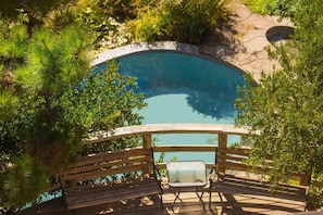 Piscina com deck de madeira e cadeiras, cercada por vegetação.