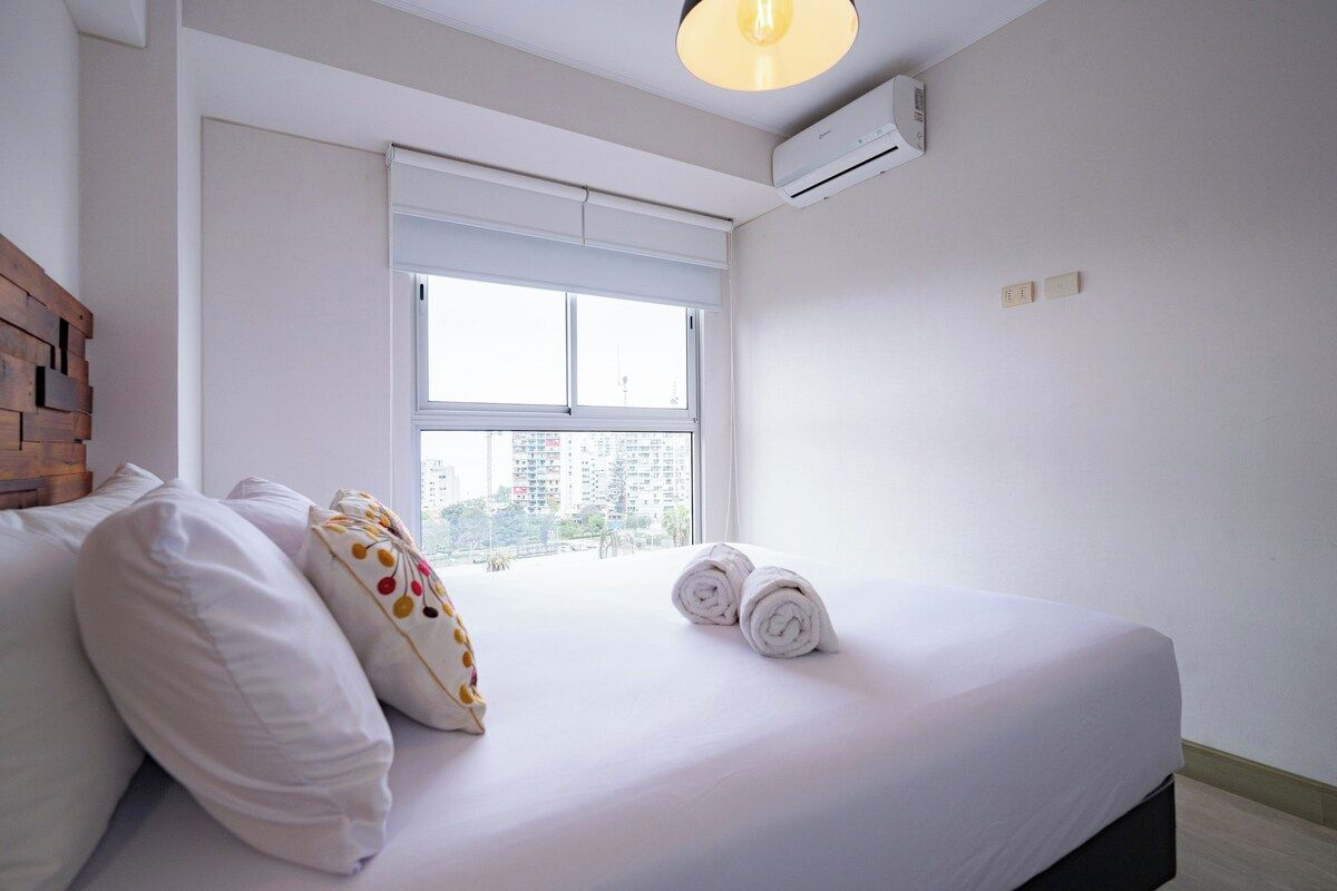 Quarto de hotel com cama, almofadas, janela e vista para prédios.