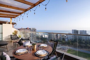 Uma área de refeições com uma mesa posta para quatro pessoas, com vista para a paisagem urbana e o mar.