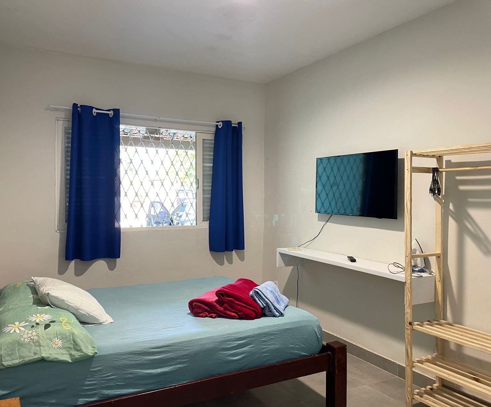 Quarto com cama, televisão na parede, janela com cortinas e prateleira de madeira.