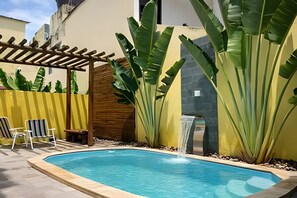 Piscina com pergolado de madeira, duas espreguiçadeiras e grandes bananeiras.