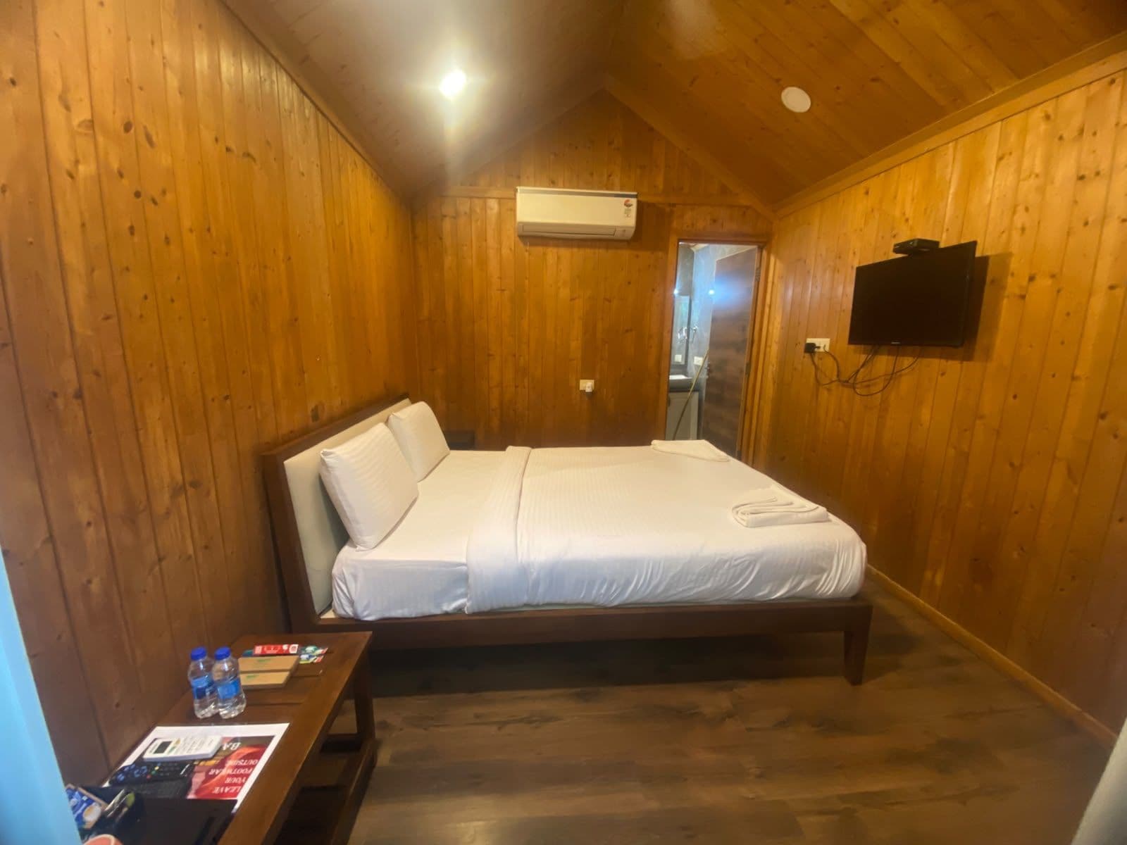 Quarto com painéis de madeira, uma cama, televisão de tela plana e uma mesinha com objetos.