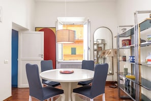 Uma área de refeições com uma mesa redonda, quatro cadeiras, um candeeiro suspenso, uma estante de livros e vista pela janela.