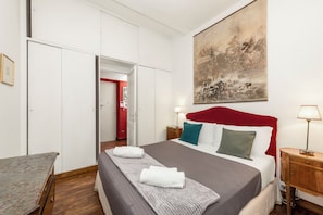 Quarto de hotel com cama, escrivaninha, cadeira, quadro na parede e porta que dá para outro cômodo.