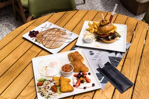 Mesa de madeira com três pratos de comida, incluindo panquecas, hambúrguer e salada com croquetes.