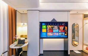 Quarto de hotel com tela digital mostrando diversos ícones de aplicativos, uma pequena mesa com um laptop, um espelho e um guarda-roupa com roupas penduradas.