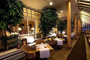 Um restaurante com mesas, cadeiras, janelas amplas e plantas em vasos.
