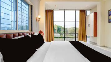 Quarto de hotel com uma cama grande, janela com cortinas, luminária fixada na parede e vista para o verde lá fora.