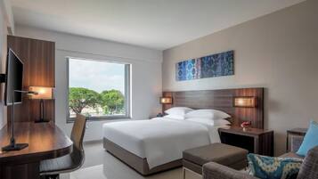 Quarto de hotel com cama, escrivaninha com cadeira, televisão e janela com vista para árvores.
