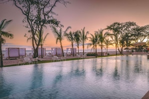 Área da piscina com espreguiçadeiras e guarda-sóis, com vista para uma praia ao pôr do sol.