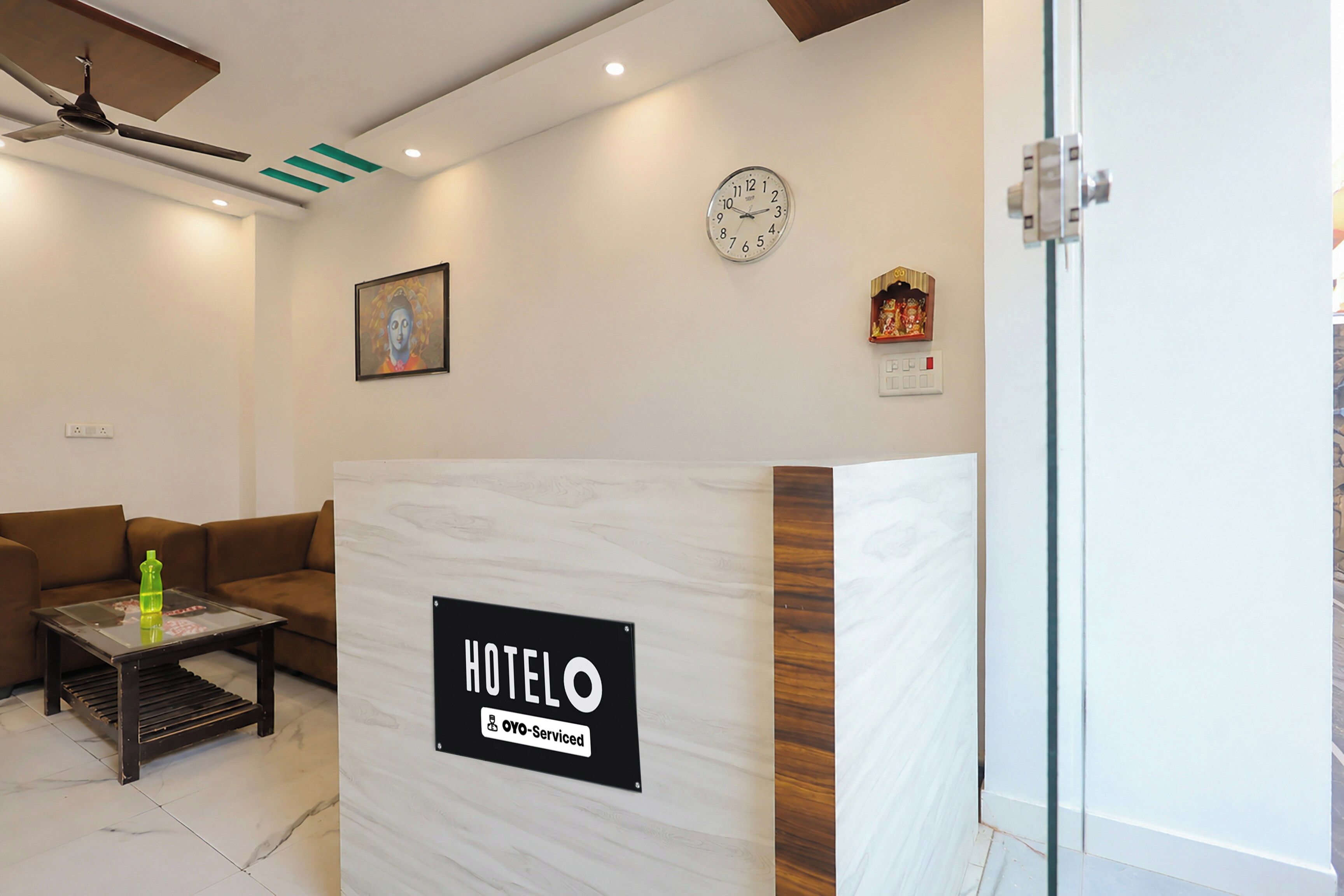 Uma área de recepção de hotel, com balcão de atendimento, relógio, quadro emoldurado e ventilador de teto.