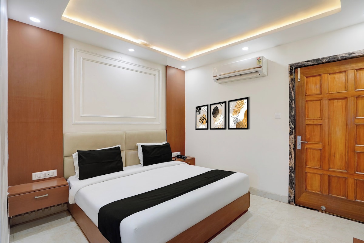 Quarto de hotel com cama, mesas de cabeceira, aparelho de ar condicionado, porta de madeira e obras de arte penduradas na parede.