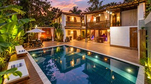 Piscina com deck de madeira, espreguiçadeiras e área de refeições com mesas e cadeiras.