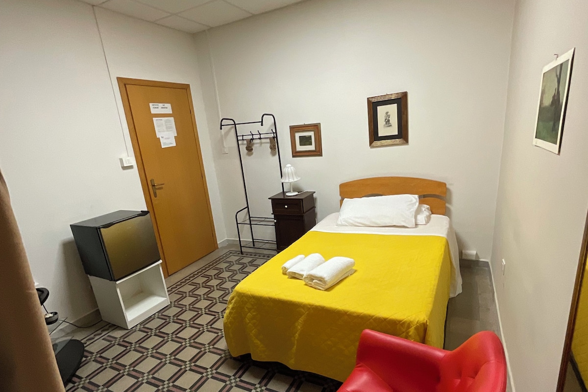 Quarto de hotel com cama, uma cadeira, escrivaninha e guarda-roupa.
