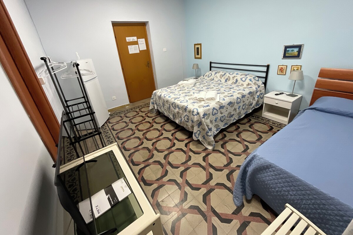 Quarto de hotel com cama, mesa de cabeceira, luminária, guarda-roupa, espelho e porta.