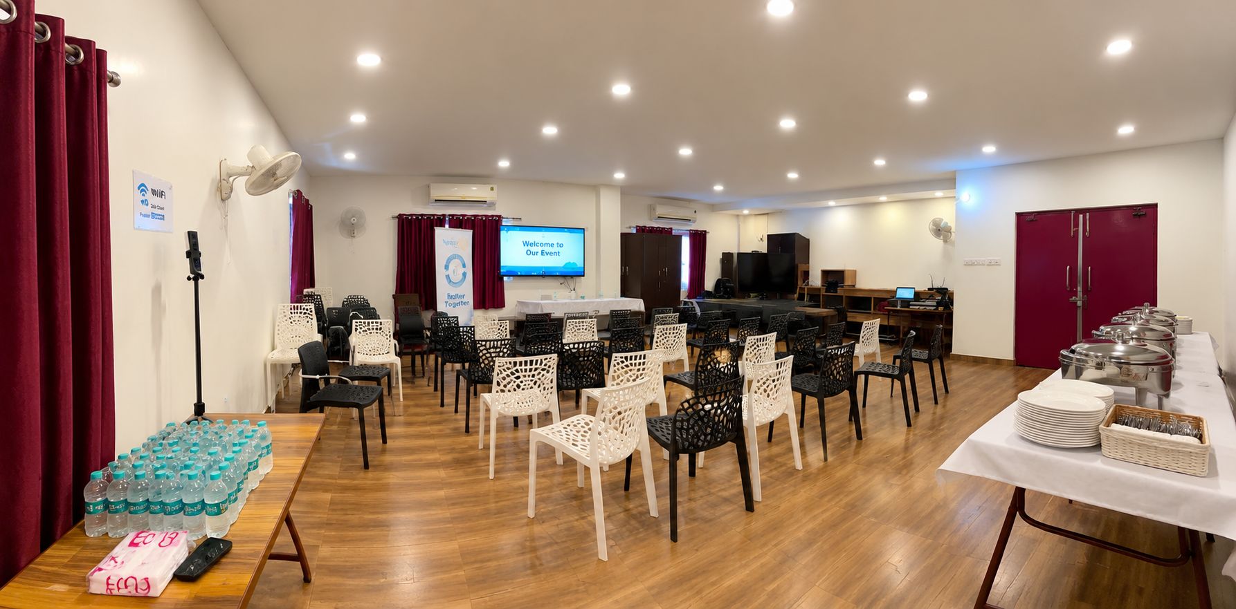 Uma sala de conferências com uma mesa branca, cadeiras preta e branca, uma tela de projetor e uma mesa de buffet com pratos e copos.