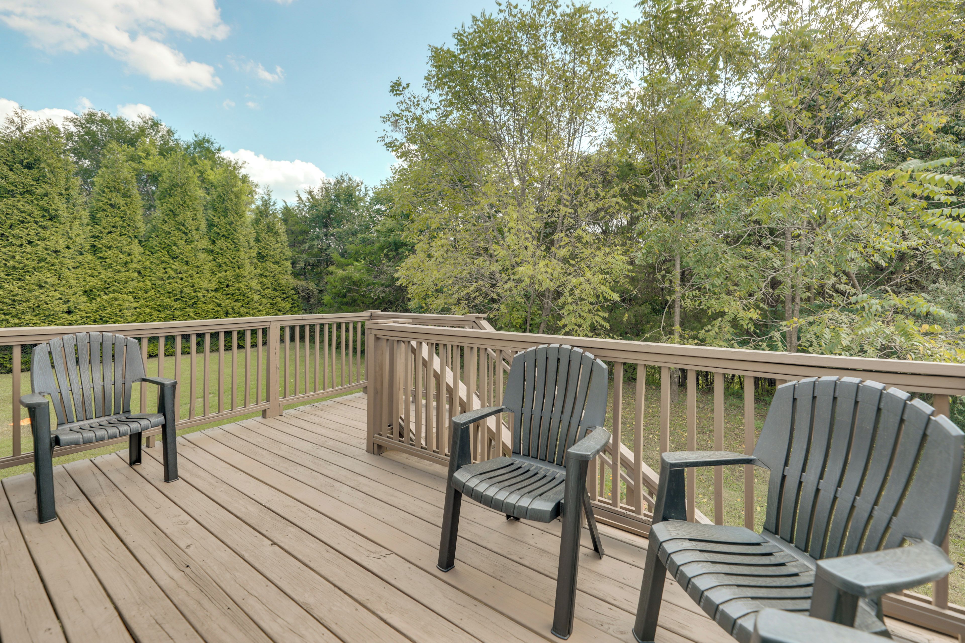 Um deck de madeira com cadeiras Adirondack e guarda-corpo com vista para uma área arborizada.