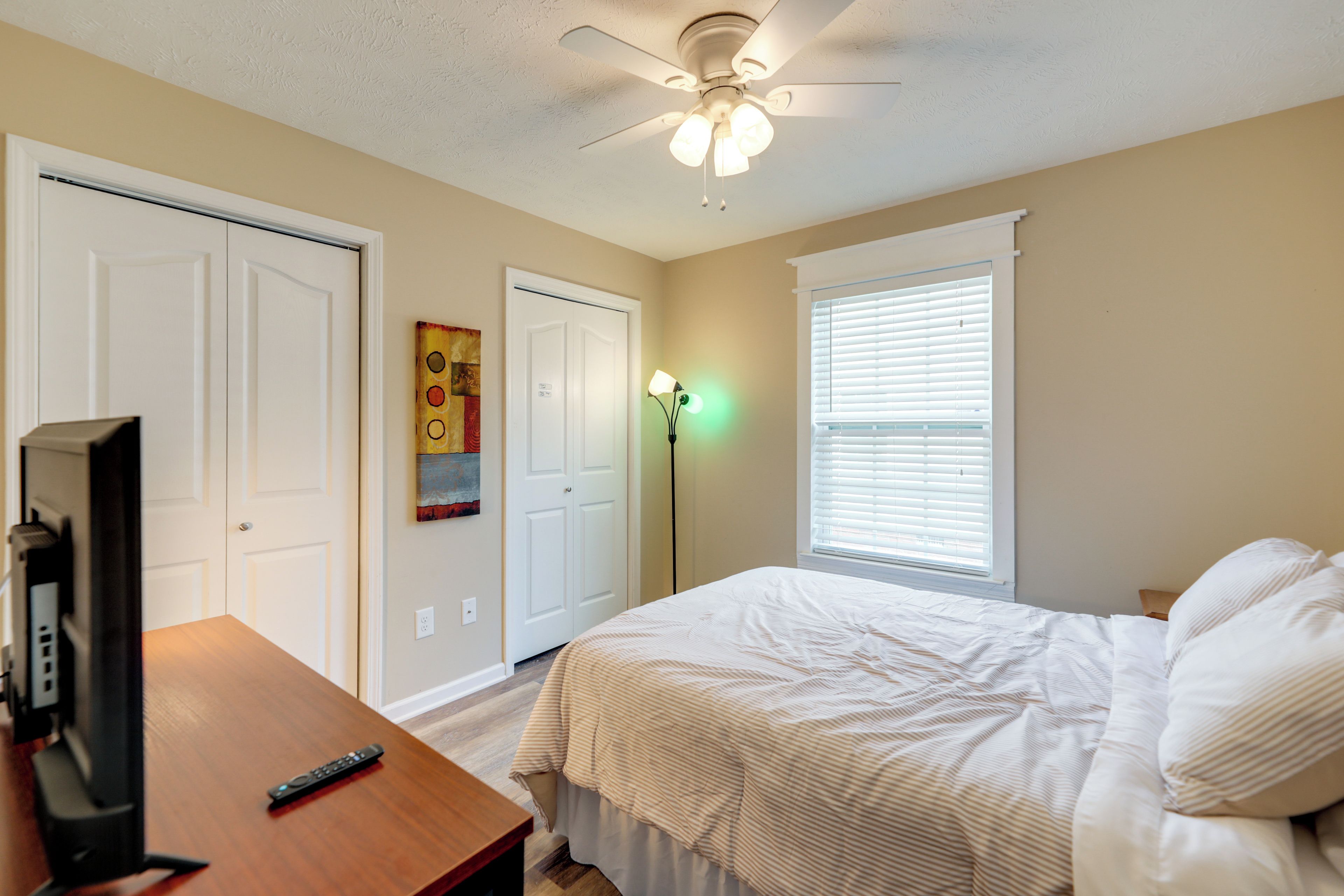 Quarto com cama, escrivaninha, televisão, ventilador de teto e janela com persianas.